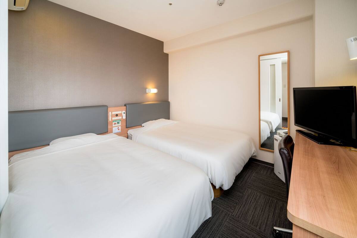 Quarto de hotel com duas camas, uma escrivaninha com televisão, um espelho e uma luminária na parede.