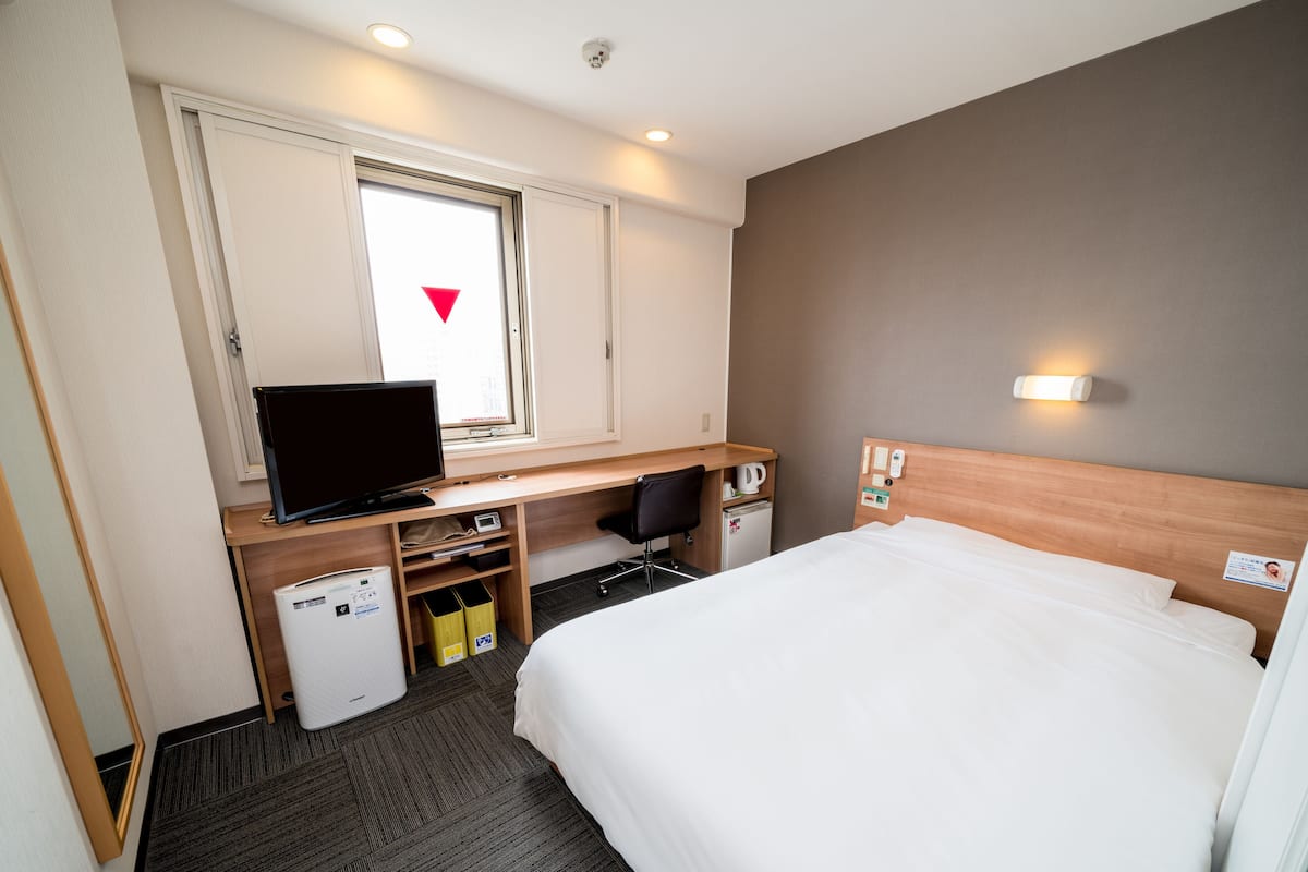 Quarto de hotel com cama, escrivaninha com computador, cadeira, pequeno frigorífico e janela com vista para edifícios.