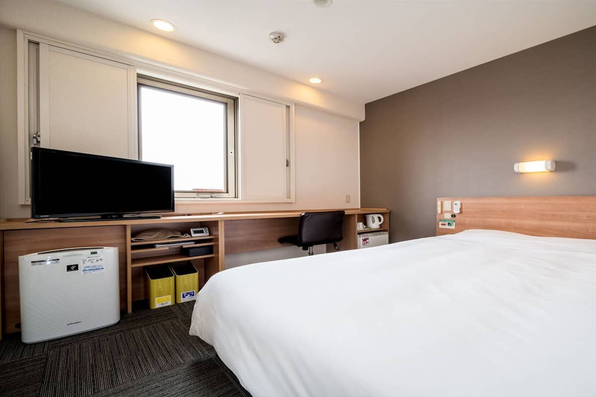 Quarto de hotel com uma cama, uma escrivaninha com televisão, um frigorífico pequeno, uma cadeira e uma janela.