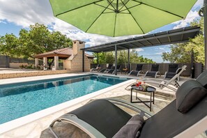 Piscina com espreguiçadeiras e mesa com bebidas, cercada por um terraço coberto.