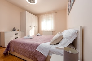 Quarto com cama, cómoda, radiador e janela com cortinas leves.
