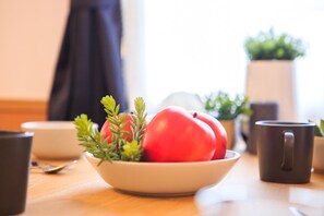 Uma tigela de maçãs vermelhas e ervas verdes sobre uma mesa de madeira, com uma caneca escura e uma tigela branca ao fundo.