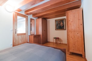 Quarto com piso de madeira, uma cama, guarda-roupa de madeira, um banquinho pequeno e um quadro na parede.