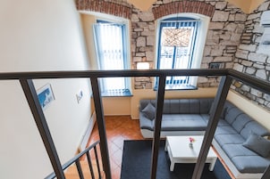 Sala de estar com parede de pedra, sofá cinzento, mesa de centro branca e vista pela janela com persianas azuis.