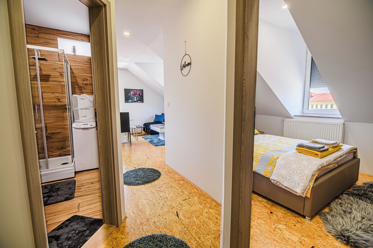 Quarto moderno com teto inclinado, cama com colcha estampada em padrão geométrico, uma pequena escrivaninha e uma cadeira.