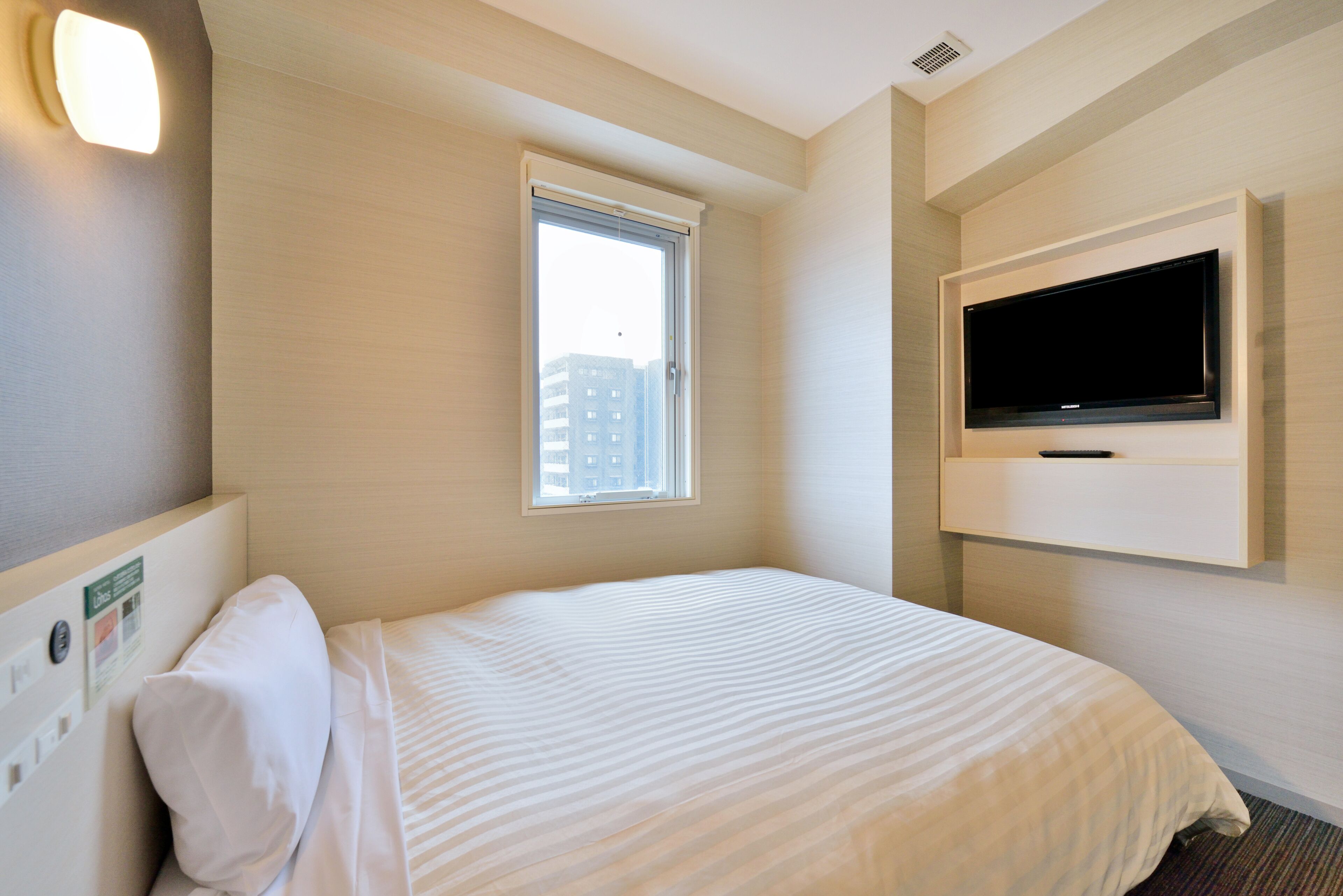 Quarto de hotel com cama, televisão presa na parede, janela e painel de controle na parede.