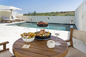 Uma área de refeições à beira da piscina, com uma mesa redonda de madeira, duas cadeiras brancas com almofadas, uma cesta de pão, um prato de frutos do mar e um copo de vinho branco.