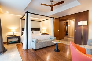 Uma cama com dossel e cortina, uma mesa de cabeceira com abajur, uma pequena mesa redonda com abajur, um sofá e piso de madeira.