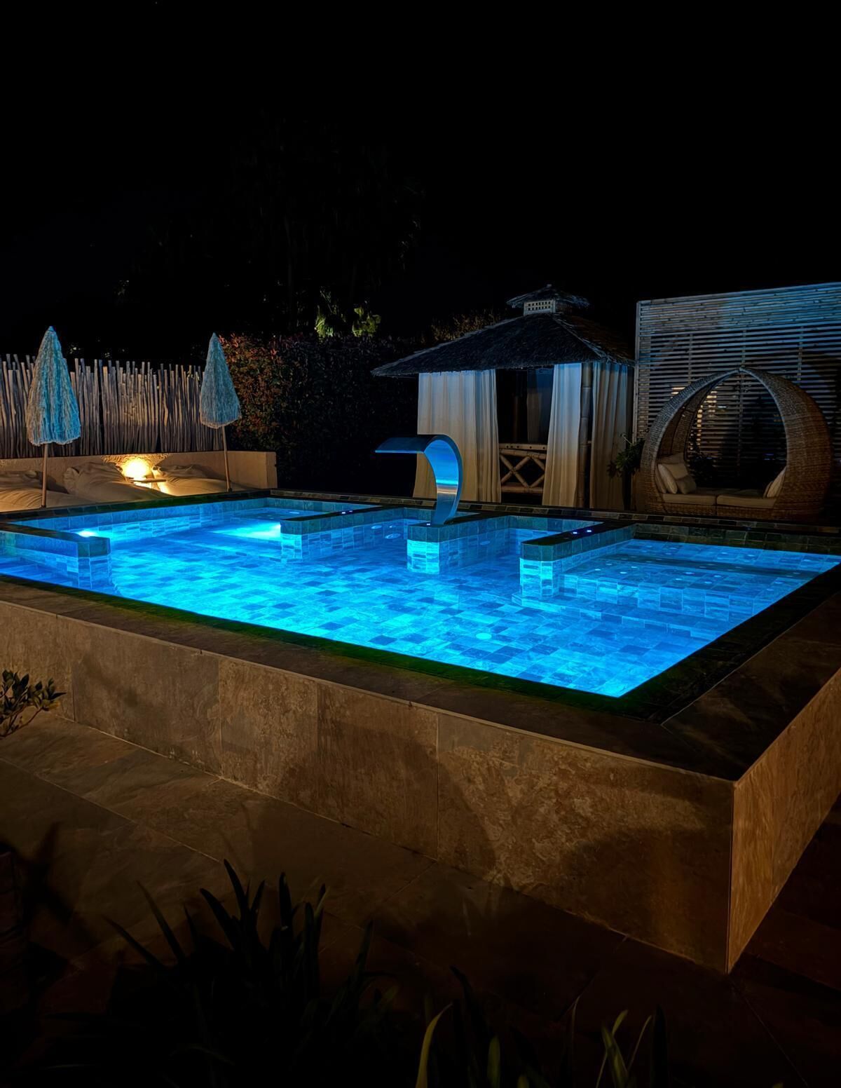 Piscina com design geométrico, ladeada por um deck de pedra. Há duas estruturas em formato de guarda-sol e um pequeno pavilhão com telhado de palha.
