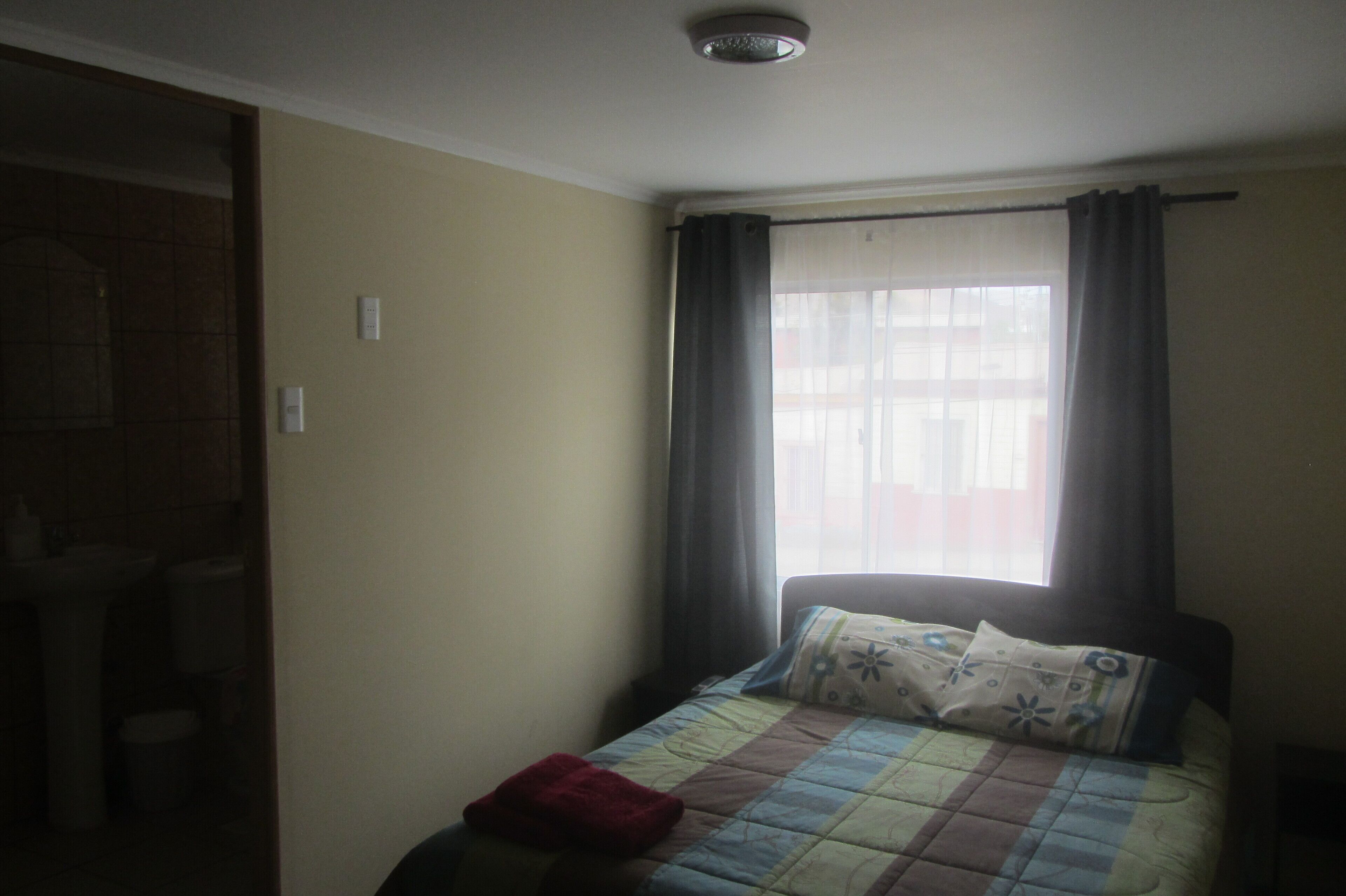 Quarto com cama, janela com cortinas e banheiro visível através de uma porta aberta.