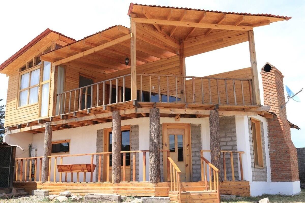 Uma casa de madeira de dois andares, com varanda, chaminé de tijolos e alpendre de madeira.