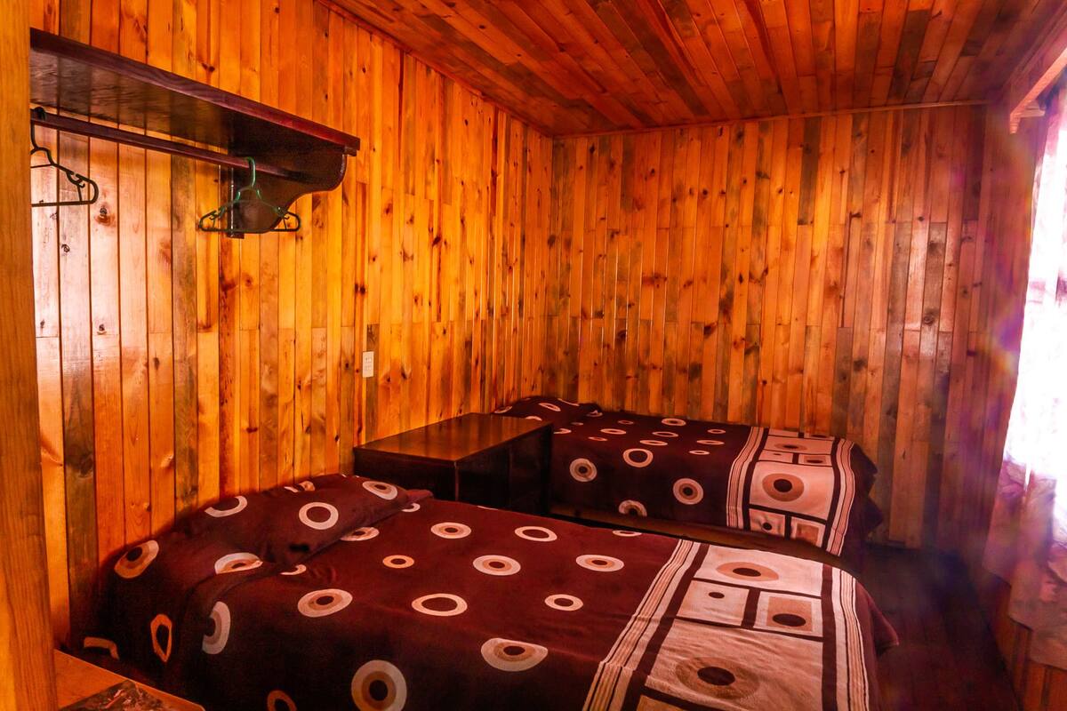 Quarto de madeira com duas camas, uma mesa e um candeeiro pendente.