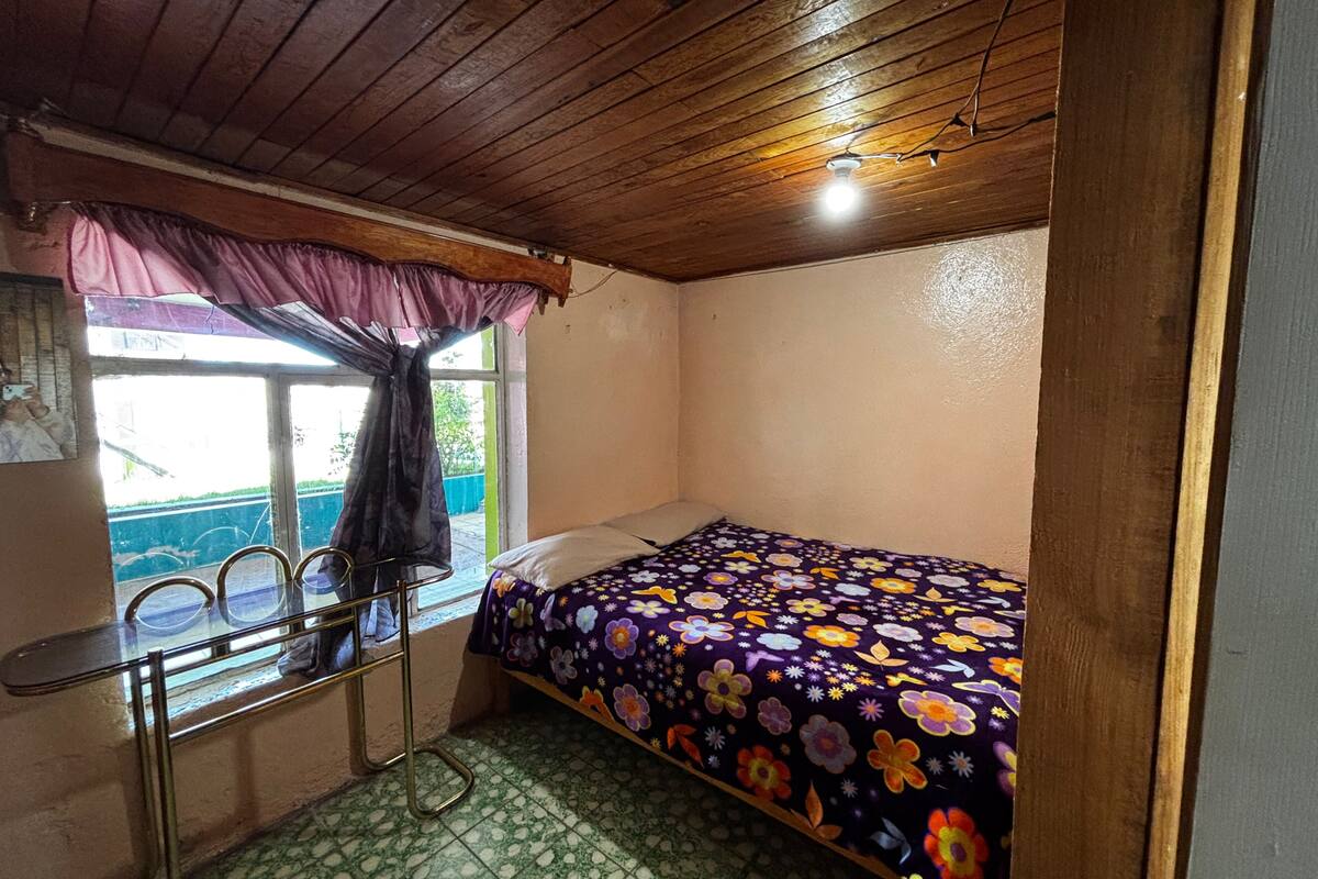Quarto pequeno com uma cama, janela com cortina, cadeira de metal e teto de madeira.