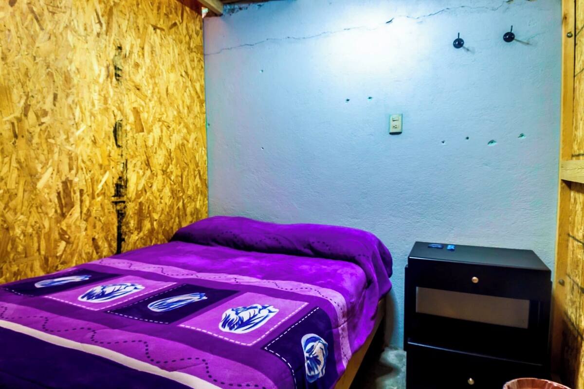 Quarto com uma cama, parede de madeira, parede branca com tomadas e mesa de cabeceira preta.