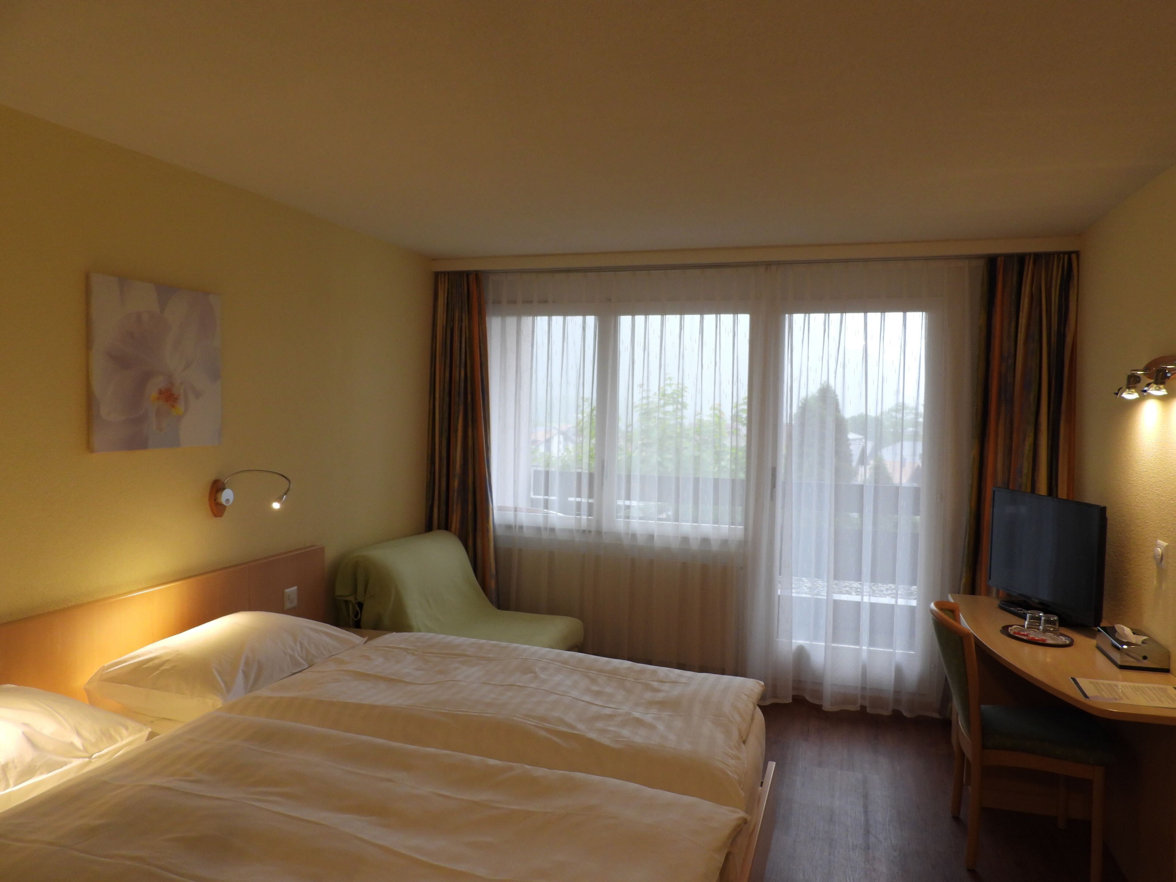 Quarto de hotel com duas camas, uma escrivaninha com cadeira, televisão e janela com cortinas.