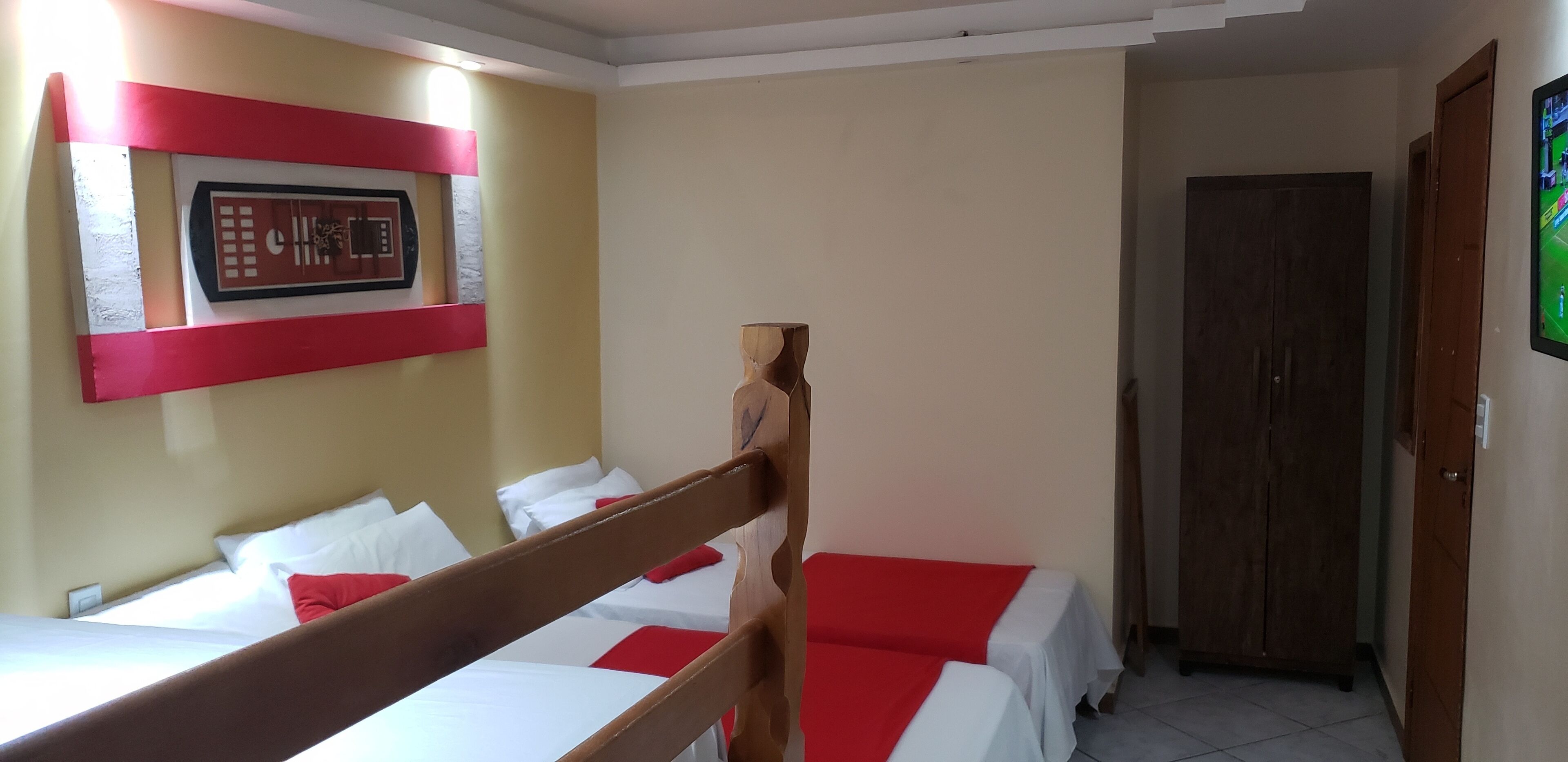 Quarto de hotel com cabeceira de madeira, duas camas de solteiro com roupa de cama vermelha e branca, um relógio de parede e uma porta de madeira.