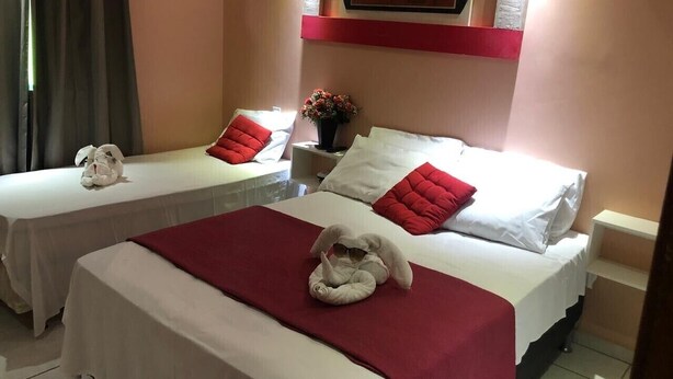 Quarto de hotel com duas camas de solteiro, cada uma com roupa de cama branca e almofadas vermelhas. Um cobertor vermelho está sobre a cama maior, e uma escultura de toalha repousa na cama menor.