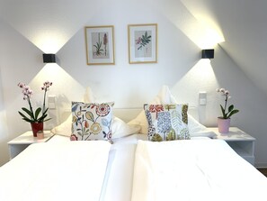 Quarto de hotel com cama, duas mesas de cabeceira, duas luminárias fixadas na parede, duas gravuras botânicas emolduradas e duas orquídeas em vasos.