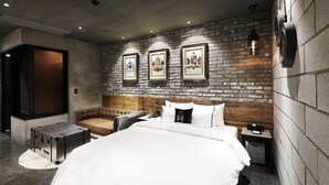 Quarto de hotel moderno com parede de tijolos, uma cama grande, cabeceira de madeira, um pequeno espaço para sentar e quadros nas paredes.