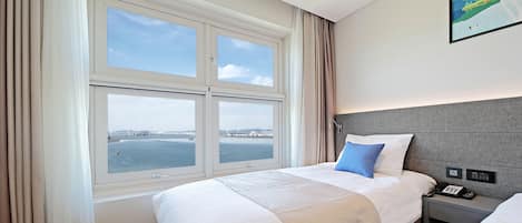 Quarto de hotel com uma janela grande, cama com roupa de cama branca, mesa de cabeceira com telefone e cabeceira cinza.