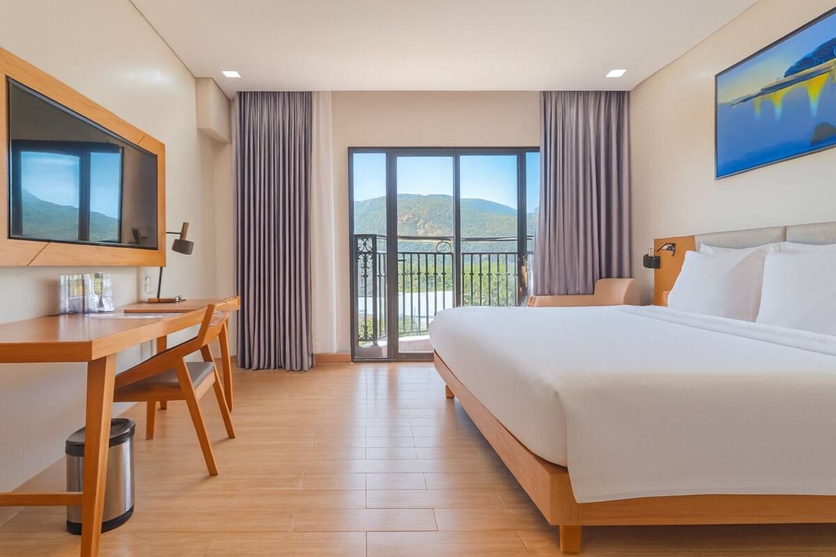 Quarto de hotel com cama, escrivaninha, cadeira, televisão e varanda com vista para as colinas.