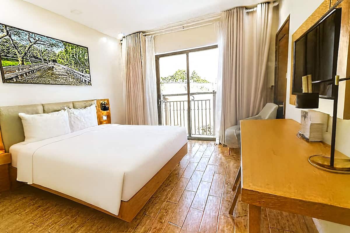 Quarto de hotel com uma cama grande, uma escrivaninha, uma cadeira, uma TV e uma varanda com cortinas.