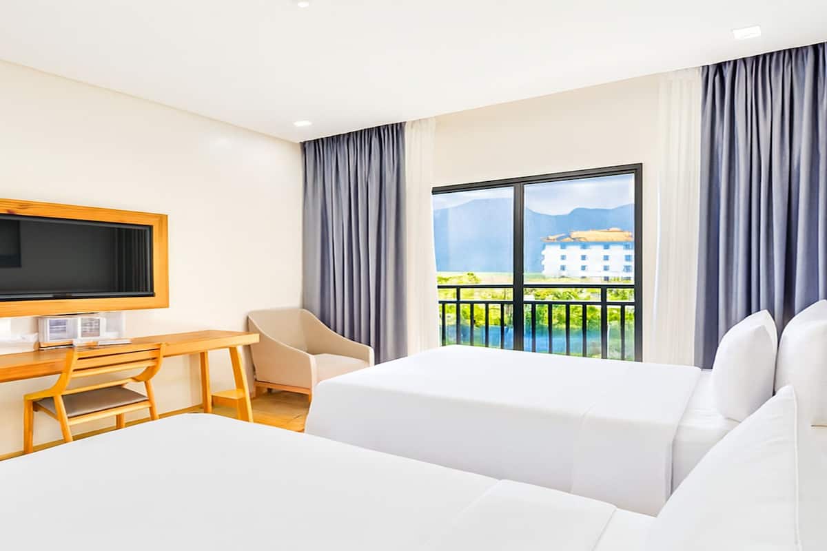 Quarto de hotel com duas camas, uma escrivaninha com televisão e vista para um edifício e vegetação pela janela.
