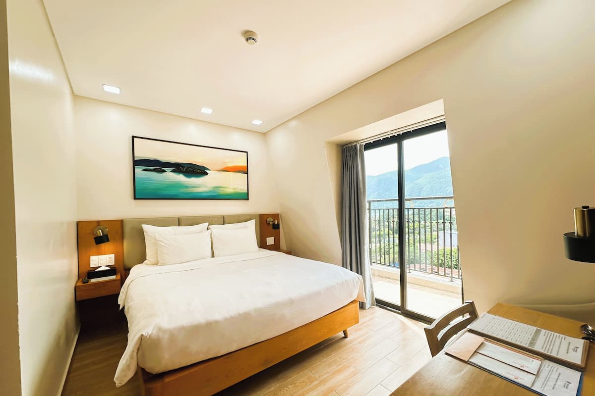 Quarto de hotel com cama, escrivaninha com papéis, um quadro e varanda com vista.