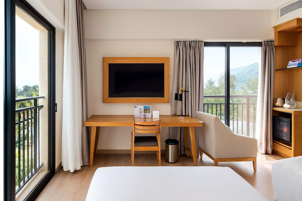 Quarto de hotel com escrivaninha de madeira, uma cadeira, uma cama, televisão e varanda com vista para as montanhas.