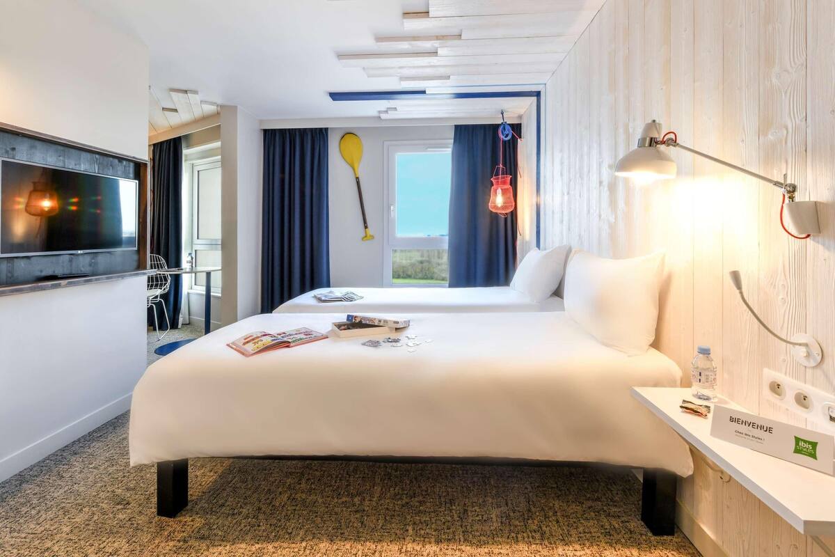Quarto de hotel com cama, escrivaninha, televisão, janela com cortinas, uma prancha de paddle e um sinal de boas-vindas.