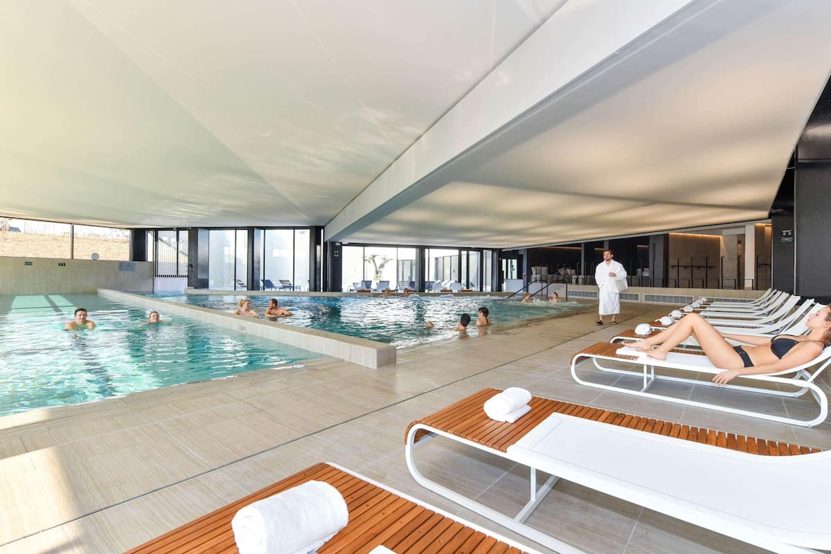 Piscina interior moderna com áreas de descanso, uma seção coberta e vista para o exterior.