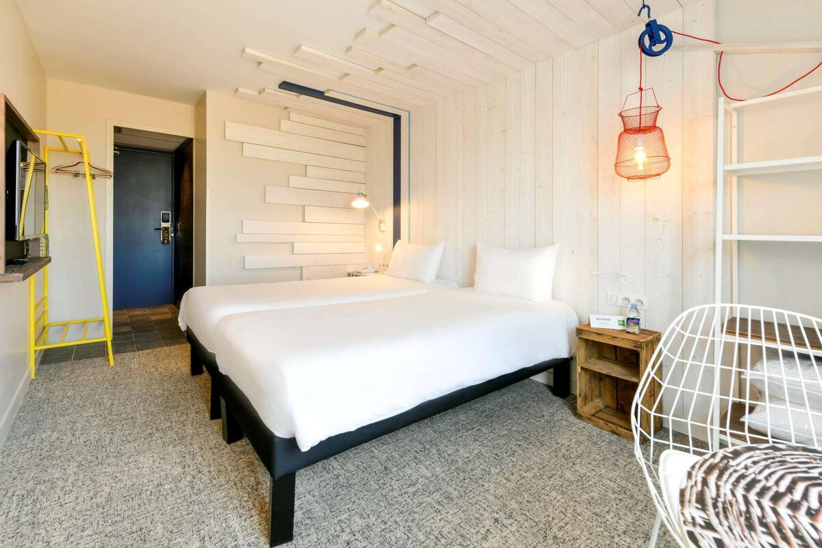 Quarto de hotel com cama, escrivaninha, cadeira, escada e prateleira fixada na parede.