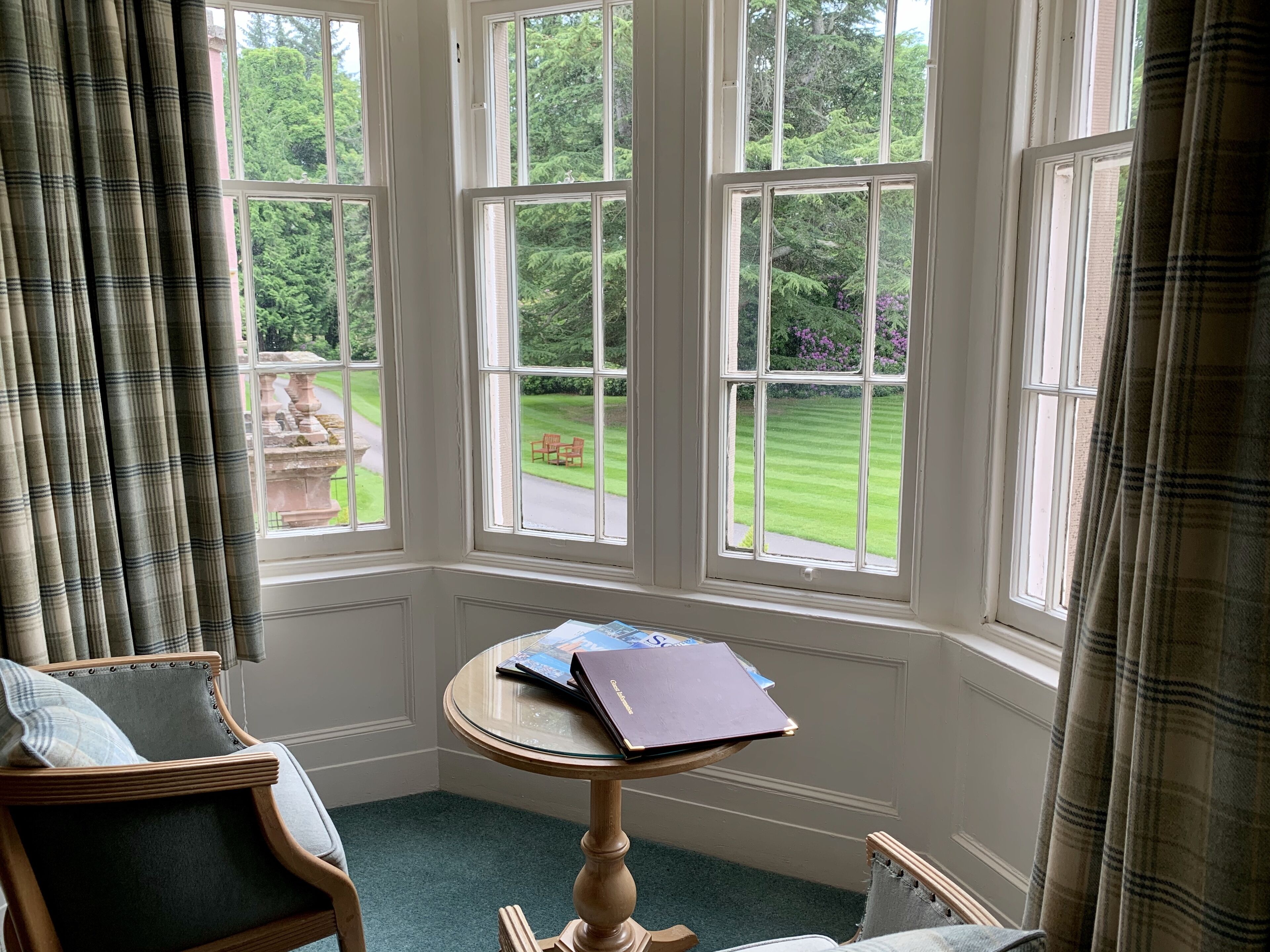 Quarto com vista para um jardim através de grandes janelas, uma pequena mesa redonda com uma pasta e uma cadeira com almofada.
