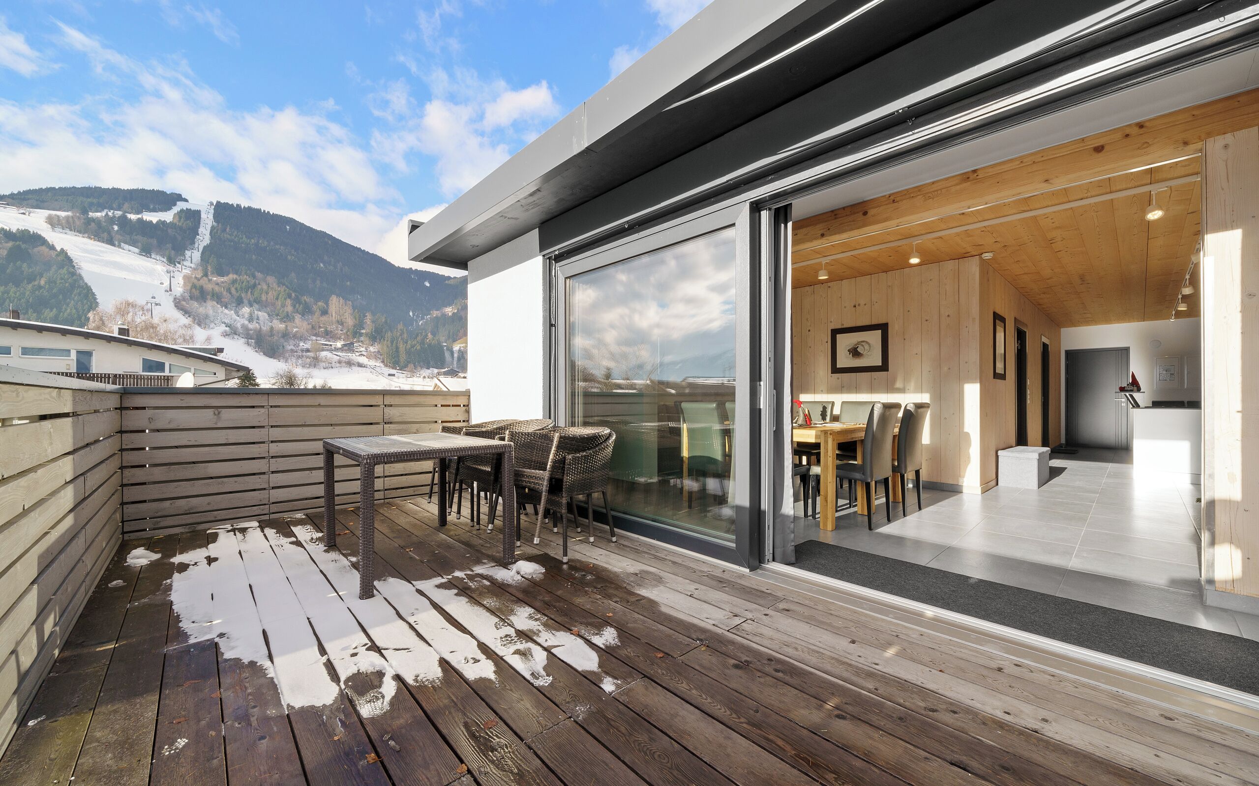 Um deck de madeira com mesa e cadeiras, uma porta de vidro que dá para um quarto com teto de madeira, e vista para uma montanha coberta de neve.
