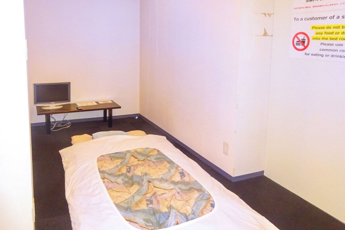 Uma cama de solteiro com colcha estampada, uma mesa de cabeceira com abajur e uma televisão sobre um suporte.