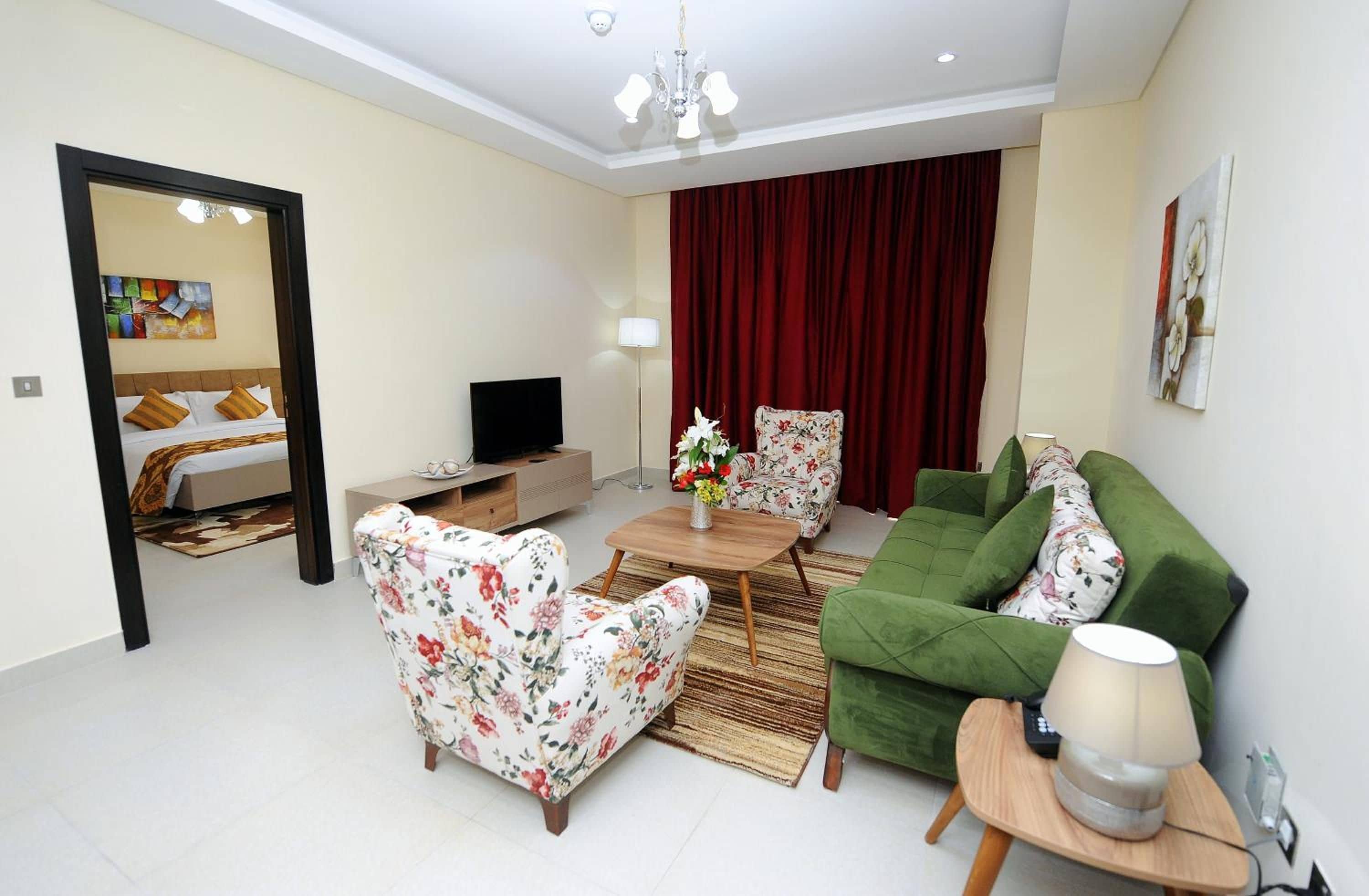 Quarto de hotel com uma cama, televisão, poltrona estampada, sofá verde e uma pequena mesa com abajur.