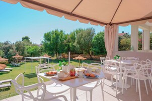 Uma área de refeições ao ar livre com cadeiras e mesas brancas, uma mesa posta com comida e bebidas, e vista para um jardim e árvores ao fundo.