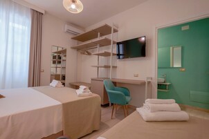 Quarto de hotel com duas camas, uma escrivaninha, uma cadeira, televisão e banheiro com parede verde.