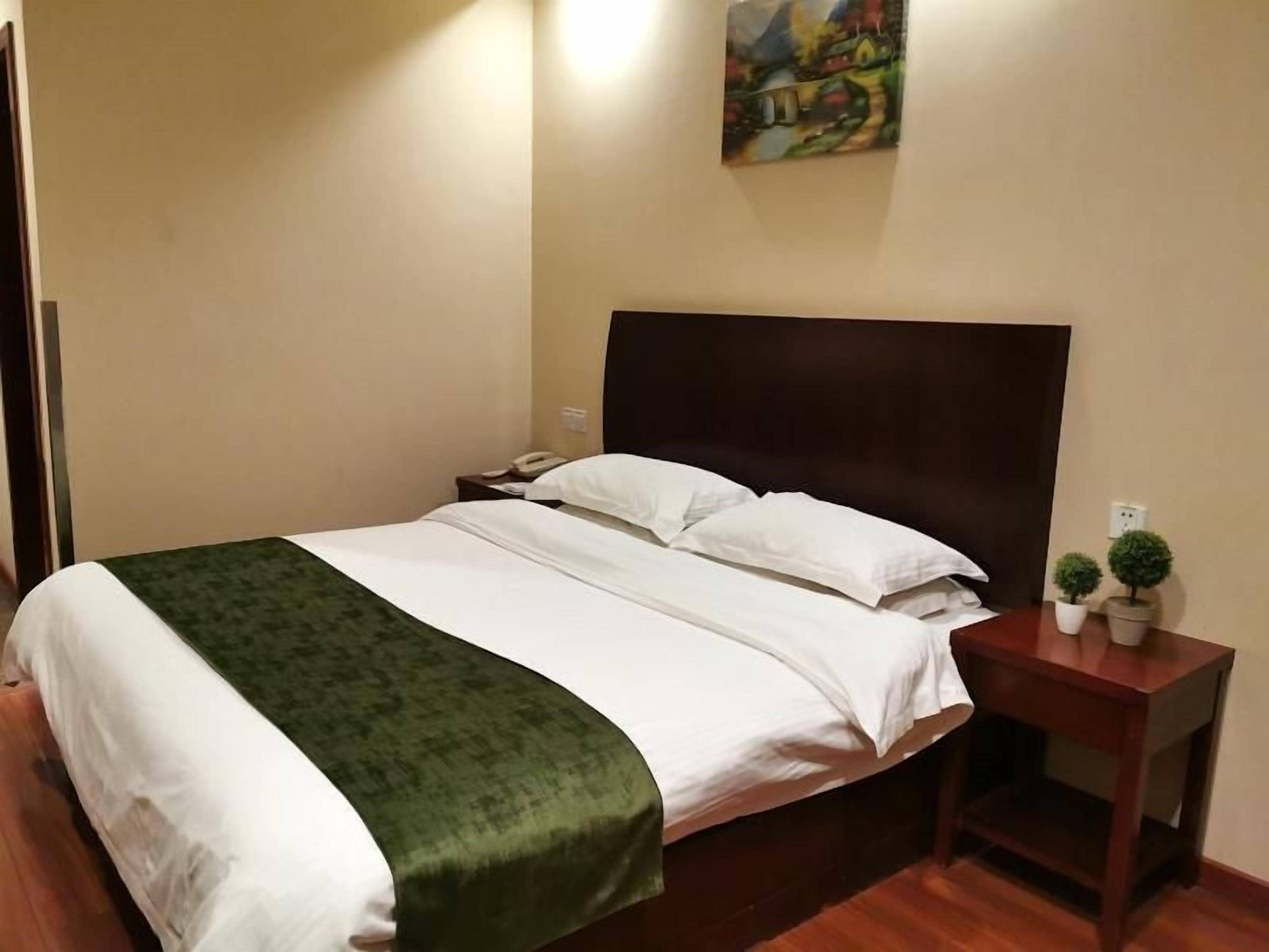 Cama bem arrumada com um manta verde, cabeceira de madeira, mesa de cabeceira com um telefone e duas pequenas plantas em vasos.