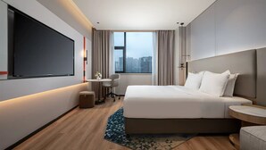 Quarto de hotel moderno com uma cama grande, televisão de tela plana, uma mesinha com um vaso e vista para o horizonte da cidade pela janela.