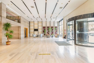 Um hall amplo com piso de madeira envernizada, balcão de recepção, área de estar e elevador de vidro.