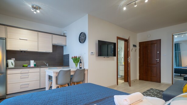 Quarto de hotel moderno com uma cama grande, uma mesa com cadeira, cozinha compacta, televisão e porta para um banheiro.