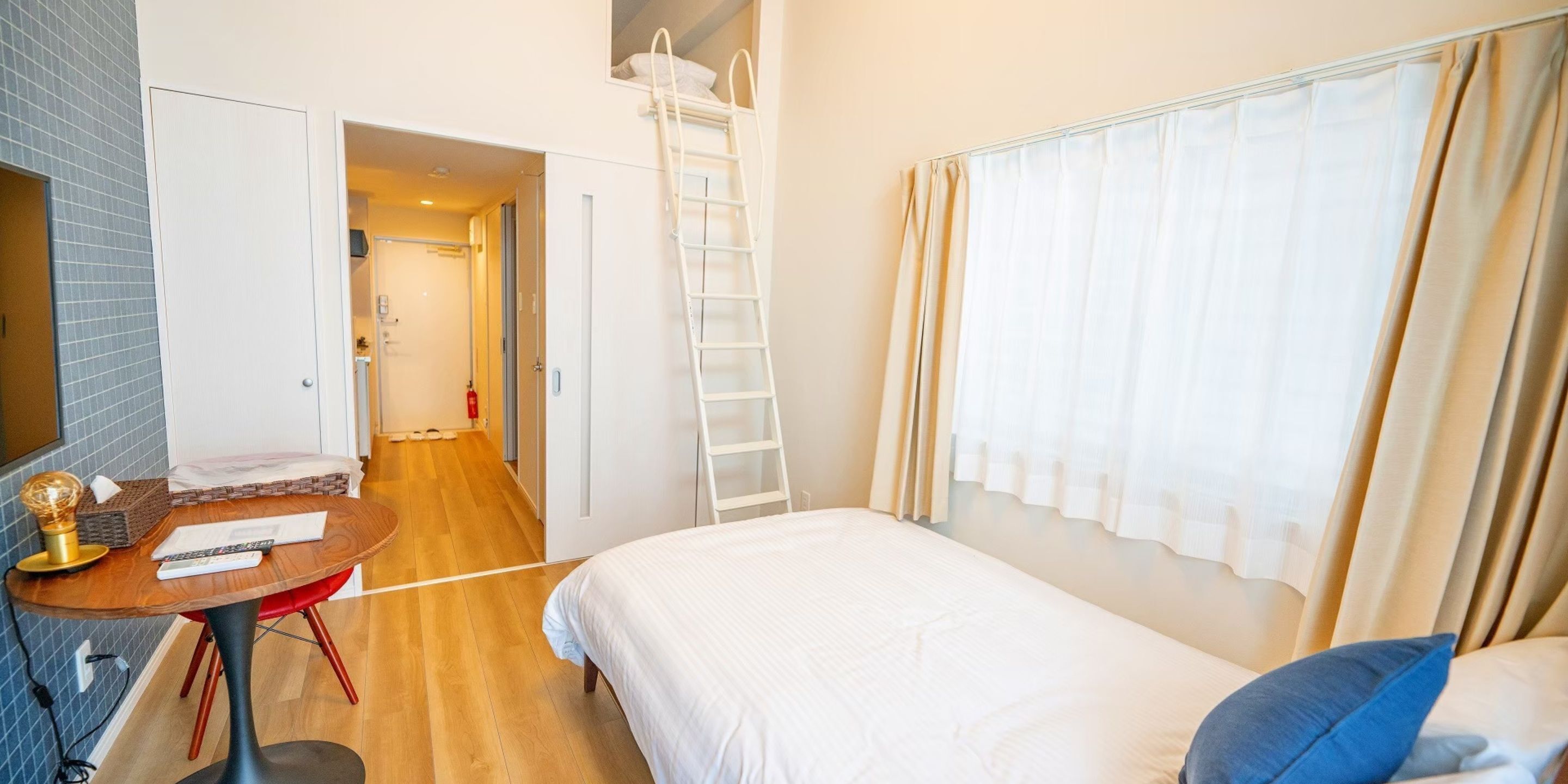 Quarto com cama, escrivaninha com abajur, escada e janela com cortinas.