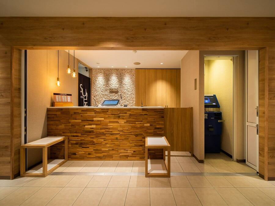 Uma área de recepção de hotel com um balcão de madeira, um painel digital e um espaço de estar pequeno.