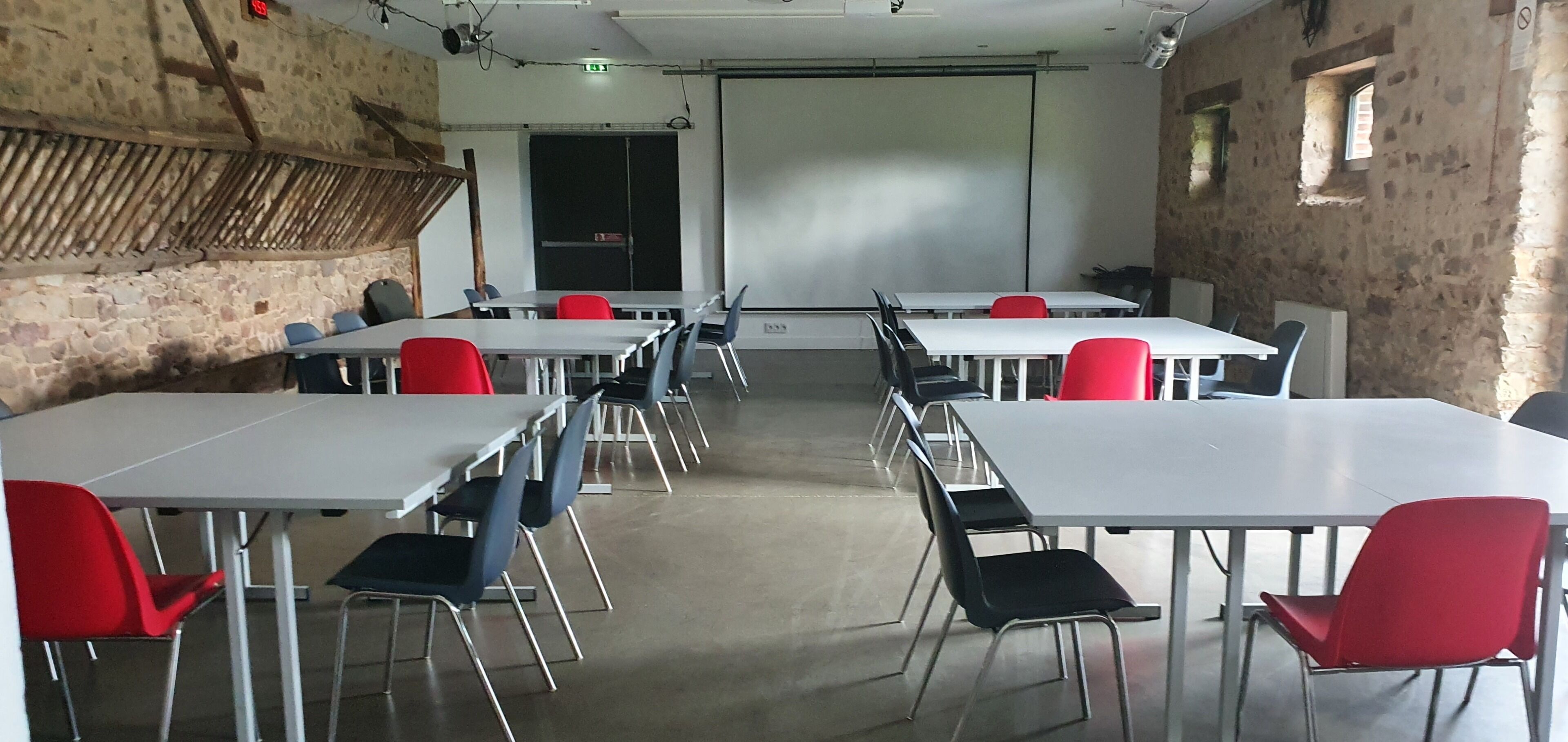 Sala de conferência com mesas brancas e cadeiras vermelhas, uma tela de projetor e paredes de pedra.