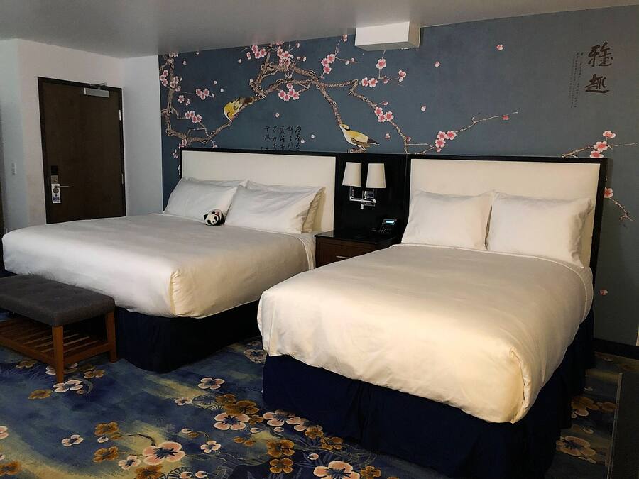 Quarto de hotel com duas camas, um banco, uma parede com arte floral e de pássaros, e uma porta.