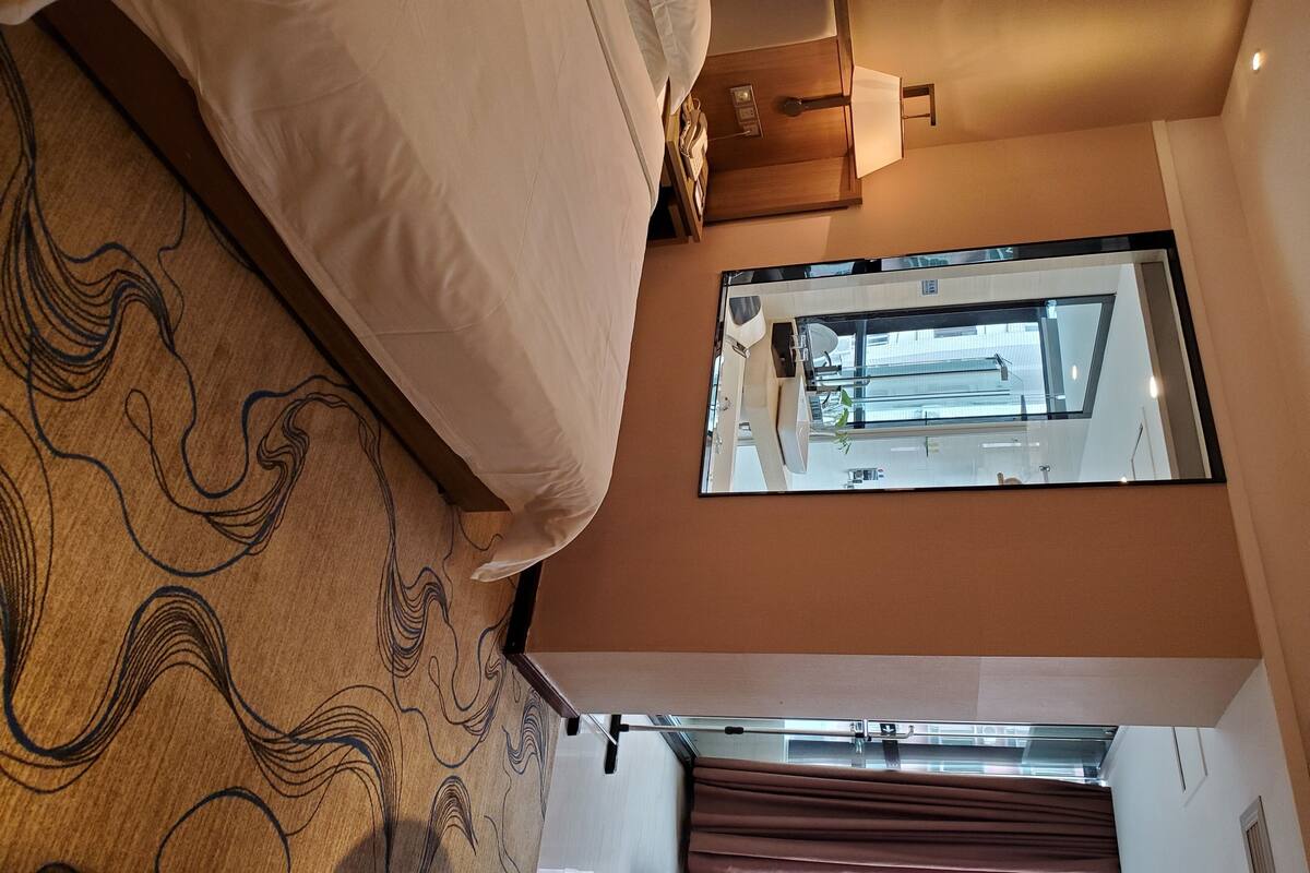 Quarto de hotel com cama, um espelho grande, janela com cortinas e tapete estampado.