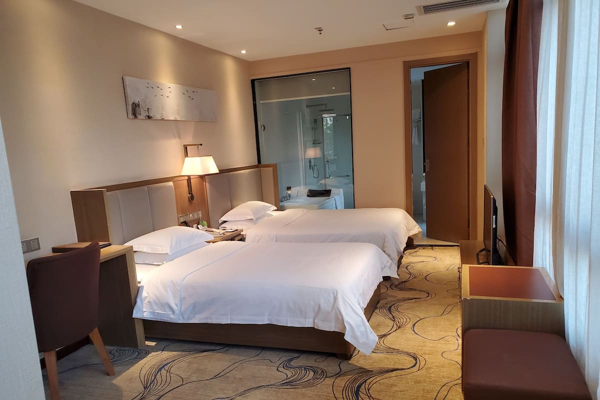 Quarto de hotel com duas camas, uma escrivaninha, uma cadeira, televisão e banheiro visível através de uma porta de vidro.