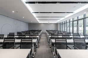 Uma sala de conferências com mesas e cadeiras dispostas em fileiras, grandes janelas e design moderno no teto.