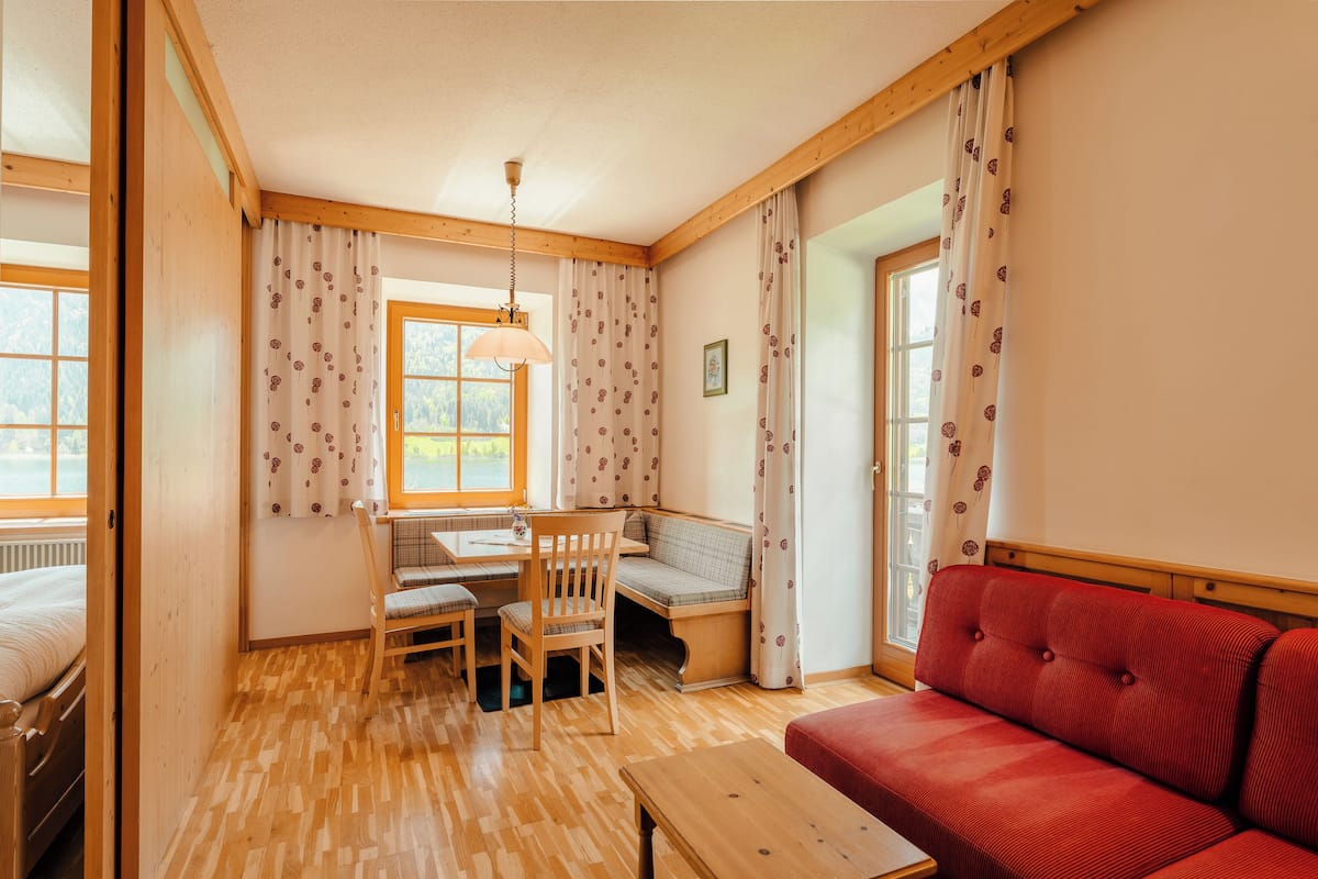 Um quarto aconchegante com piso de madeira, um sofá vermelho, um banco, uma mesa e uma janela com cortinas.